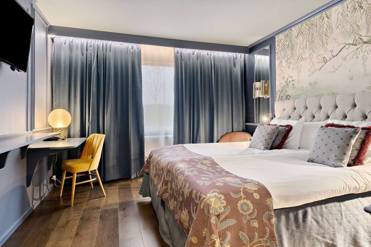 Quarto de hotel com uma cama grande, uma escrivaninha com candeeiro, uma cadeira e vista para o exterior pela janela.