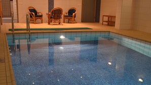 Piscina interna com chão de azulejos azuis, cadeiras de madeira e parede revestida.