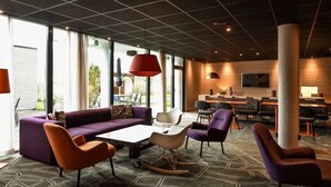 Um hall de hotel moderno com um sofá roxo, cadeiras laranja, uma mesa branca e uma luminária pendente vermelha.