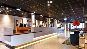 Uma área de recepção de hotel moderna, com um balcão de madeira clara, uma tela digital e um bar ao fundo.