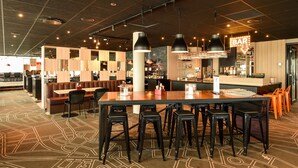 Uma área de bar moderna com uma mesa de madeira comprida, banquetas de bar pretas e um letreiro de neon com a palavra 'BAR'.