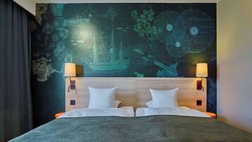 Quarto de hotel com uma cama grande, duas luminárias de cabeceira e um mural na parede com elementos náuticos e botânicos.