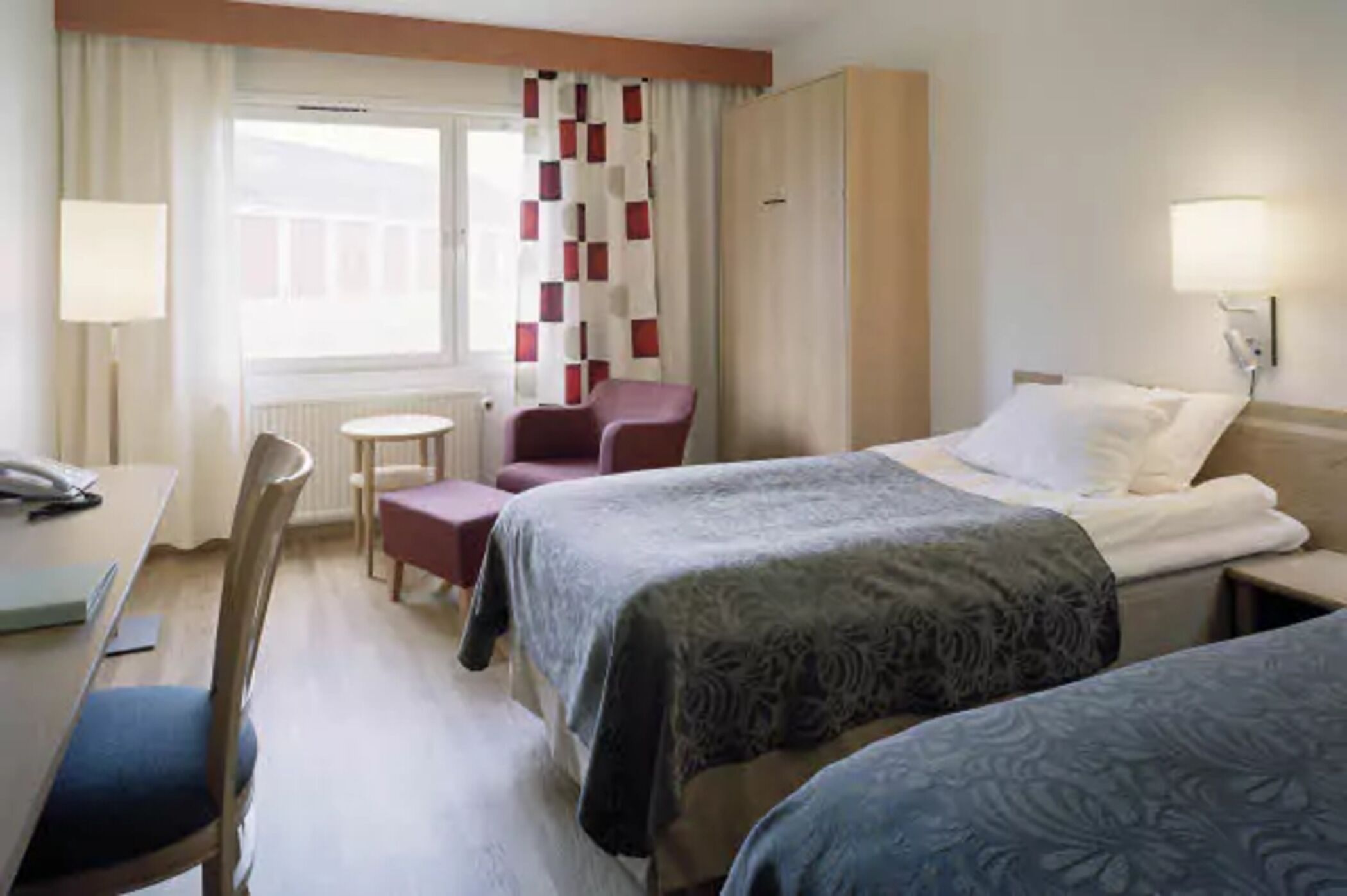 Quarto de hotel com duas camas, uma escrivaninha, uma cadeira e uma mesa pequena.