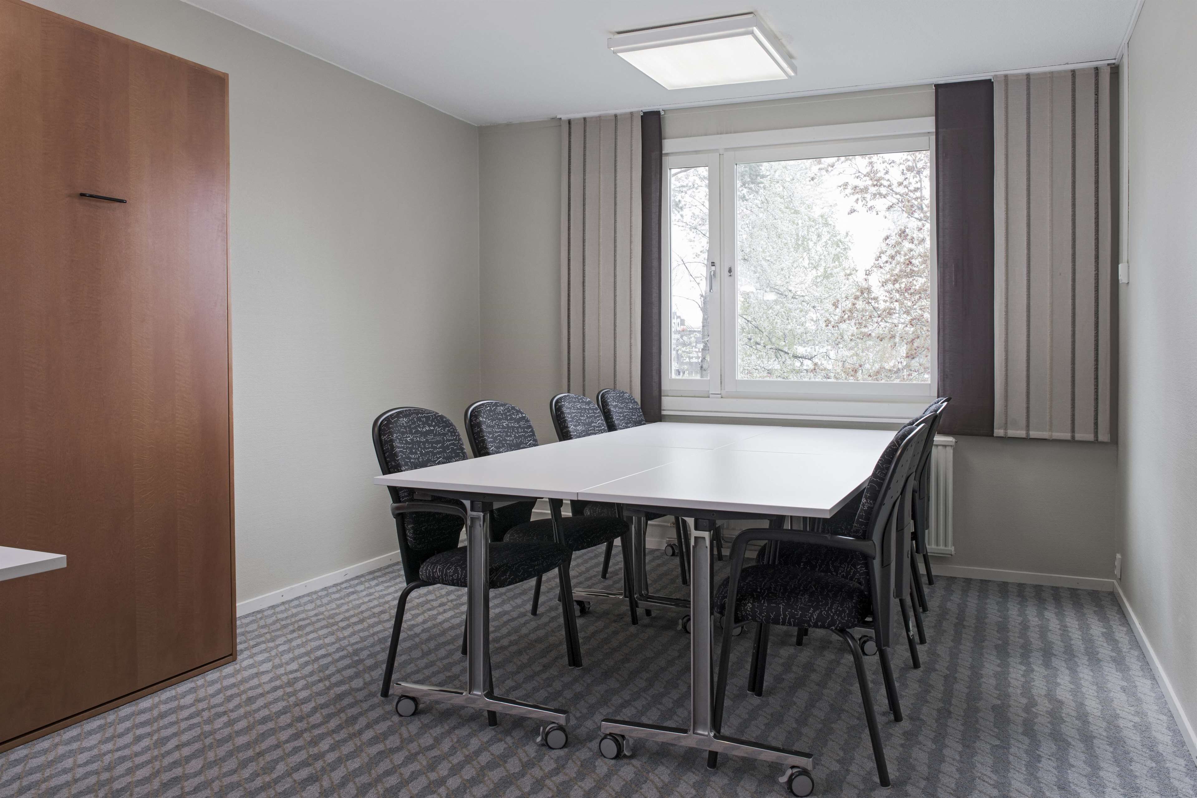 Uma sala de conferências com uma mesa grande, seis cadeiras, uma porta de madeira, uma janela com cortinas e um radiador.