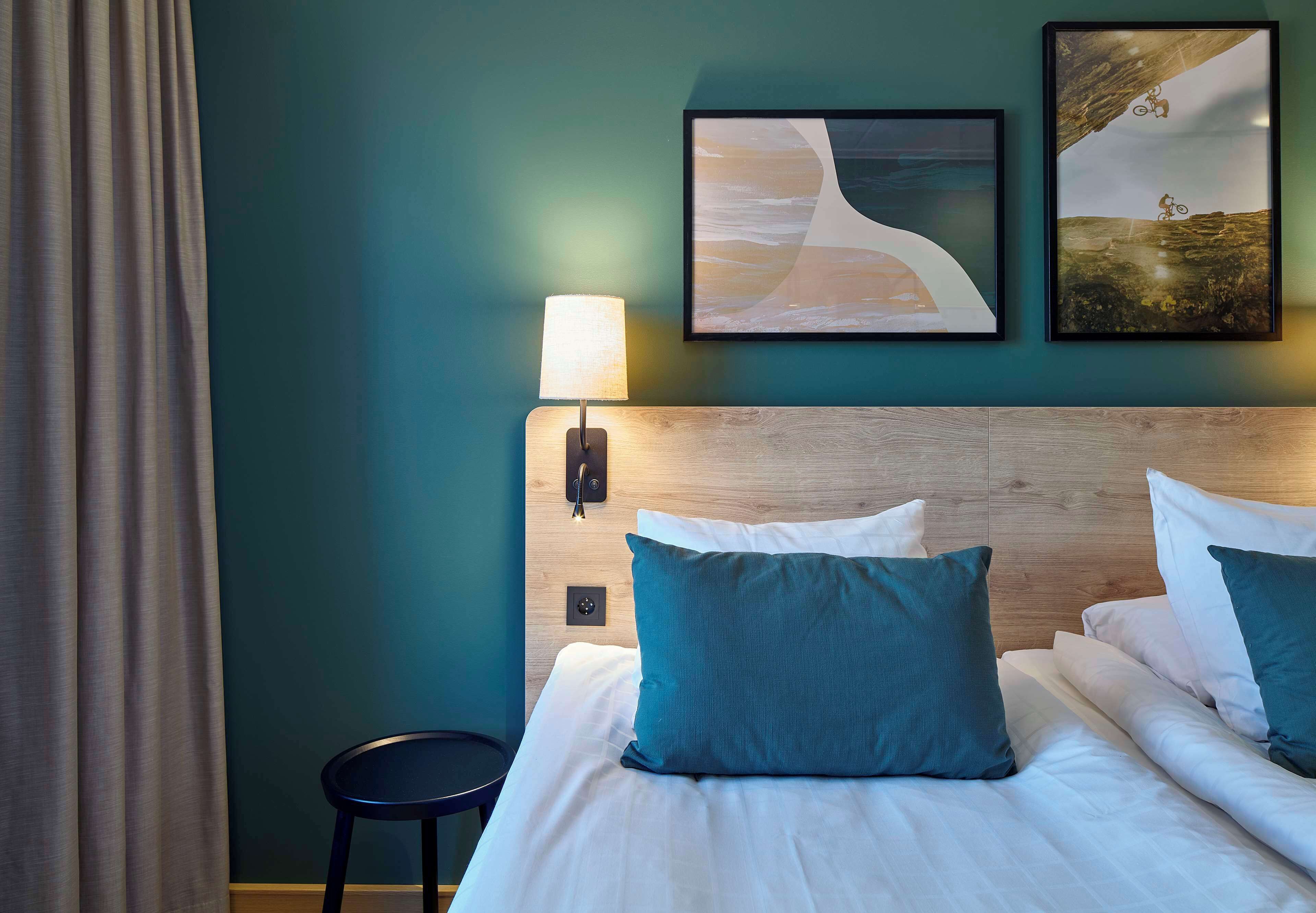 Quarto de hotel com uma cama, uma mesa de cabeceira, uma luminária e duas fotografias emolduradas na parede.