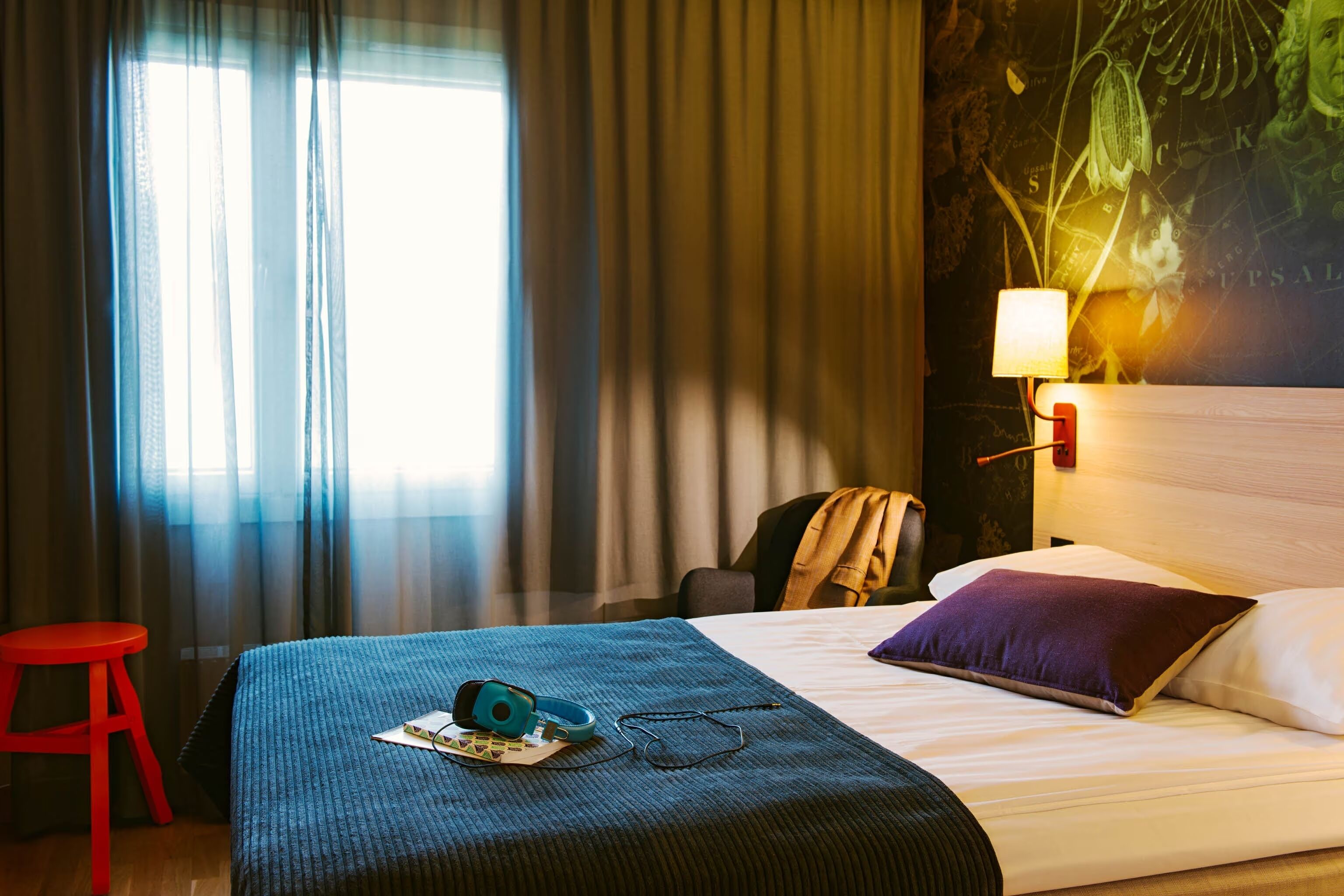 Quarto de hotel com uma cama, um banquinho vermelho, uma luminária e uma parede com um mural botânico.