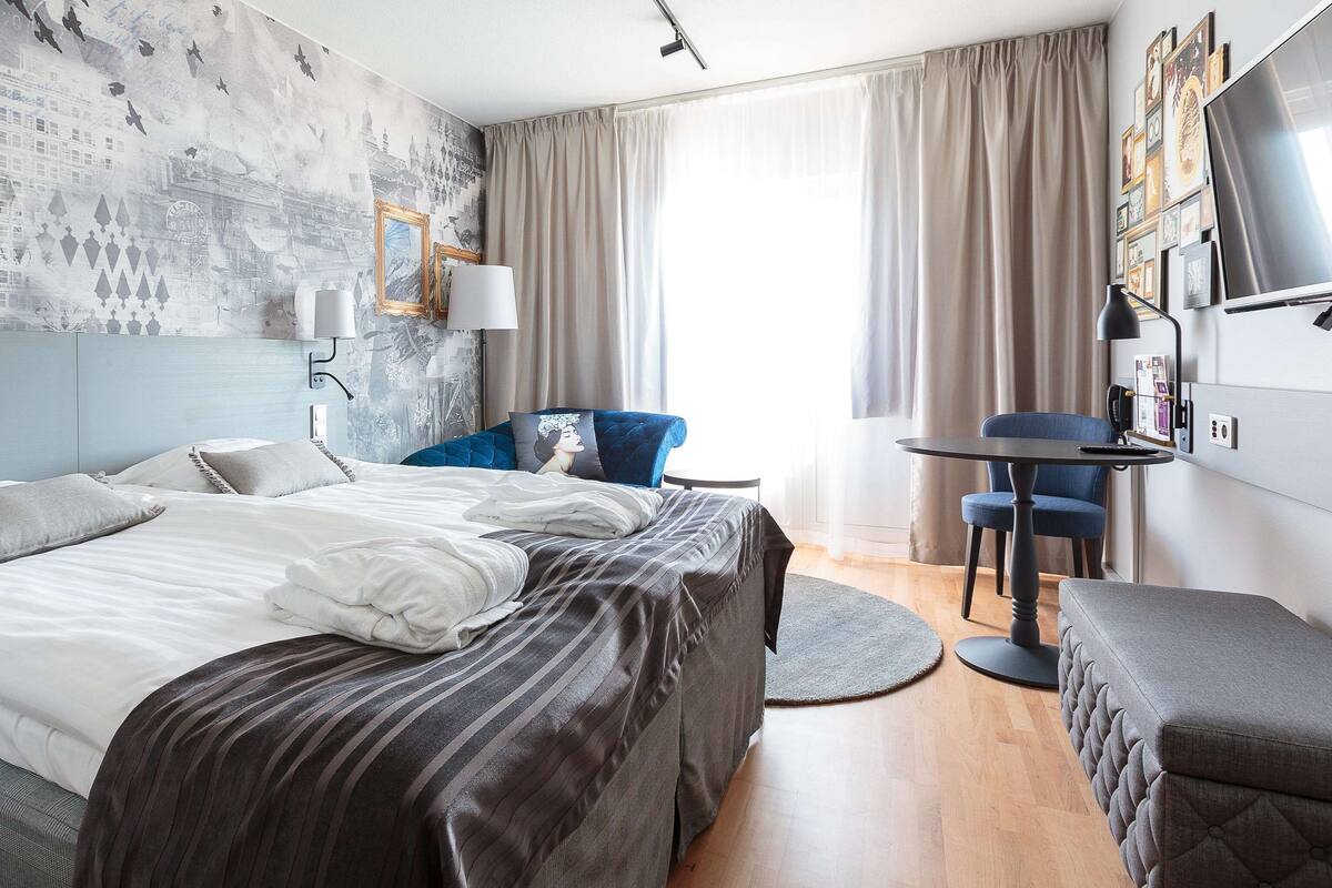 Quarto de hotel com uma cama grande, uma cadeira, uma mesa pequena e uma TV fixada na parede.