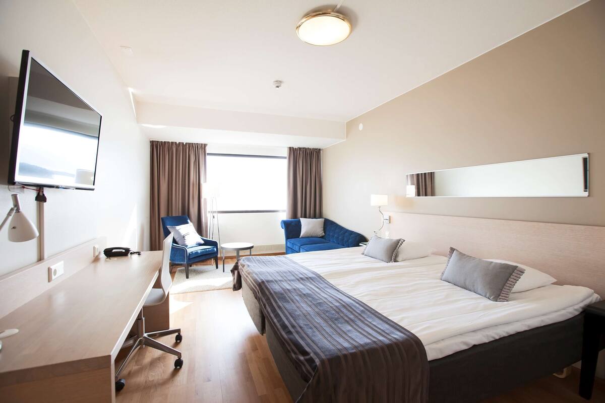 Quarto de hotel com uma cama grande, uma escrivaninha, uma cadeira, uma televisão e uma janela com cortinas.