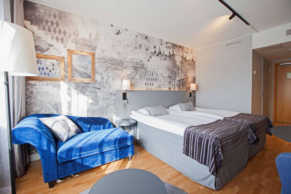 Quarto de hotel moderno com cama, sofá azul, pequena mesa e parede com mural de paisagem urbana.
