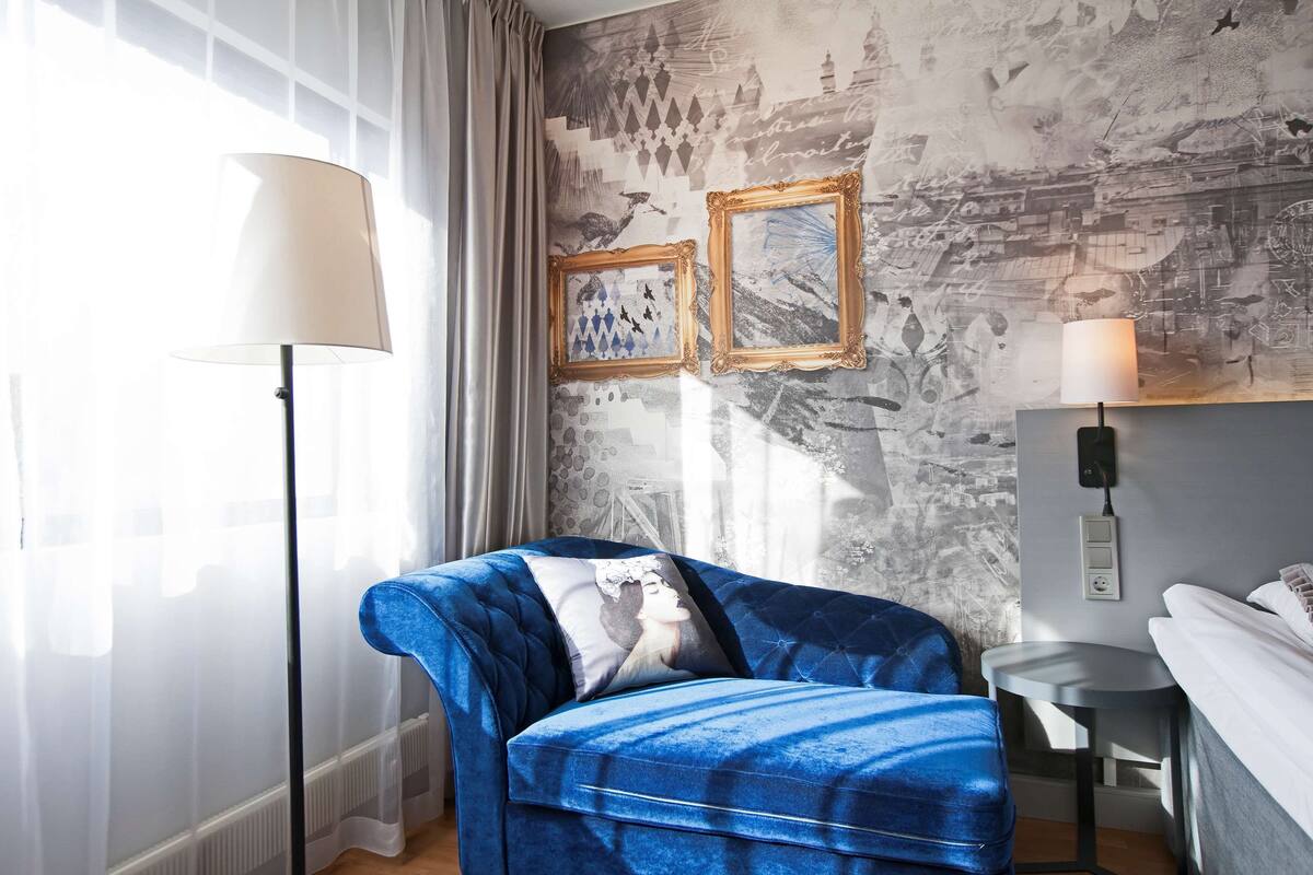 Quarto com uma cadeira estofada azul, uma cama, um abajur e uma parede com um mural da paisagem urbana.