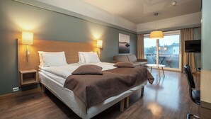 Quarto de hotel com uma cama grande, duas mesas de cabeceira com luminárias, uma pequena mesa com uma cadeira, uma TV e uma varanda com vista.