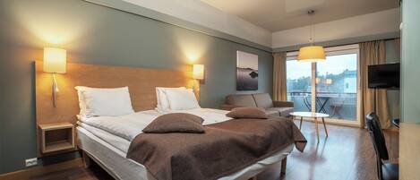 Quarto de hotel com uma cama grande, duas mesas de cabeceira com luminárias, uma pequena mesa com uma cadeira, uma TV e uma varanda com vista.