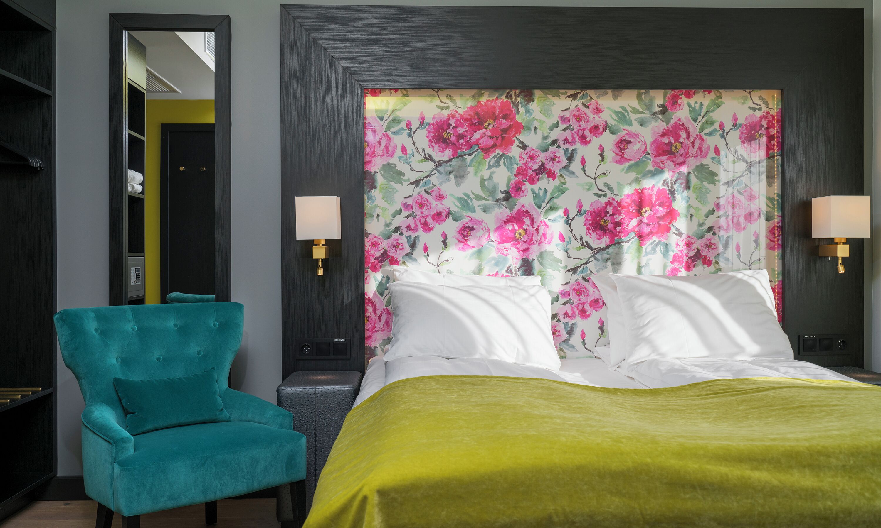 Quarto de hotel com uma cama, uma poltrona azul-esverdeada, uma mesinha de cabeceira e um espelho.