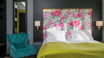 Quarto de hotel com uma cama, uma poltrona azul-esverdeada, uma mesinha de cabeceira e um espelho.