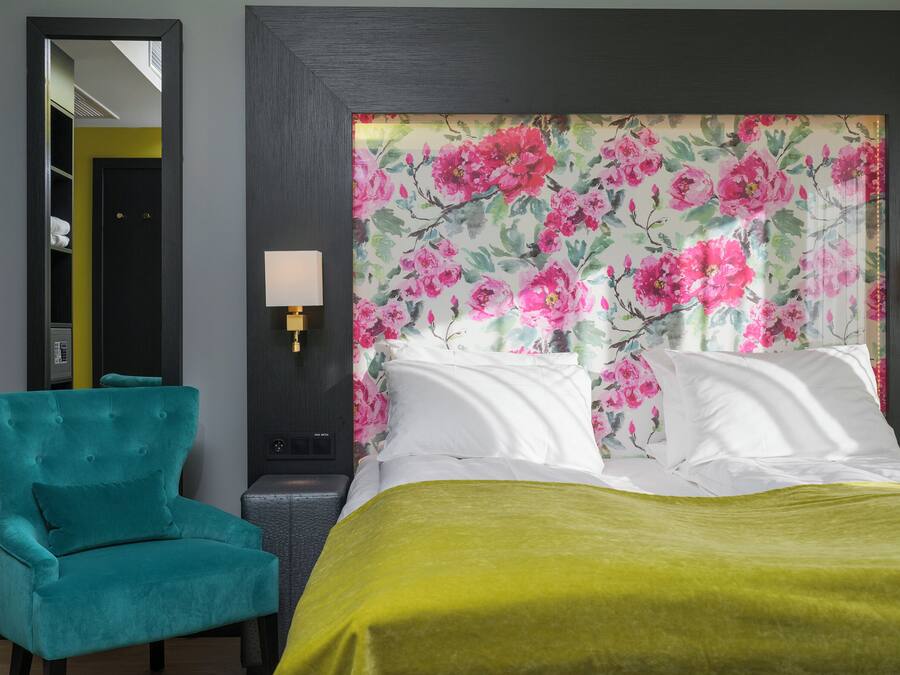 Quarto de hotel com uma cama, uma poltrona azul-esverdeada, uma mesinha de cabeceira e um espelho.