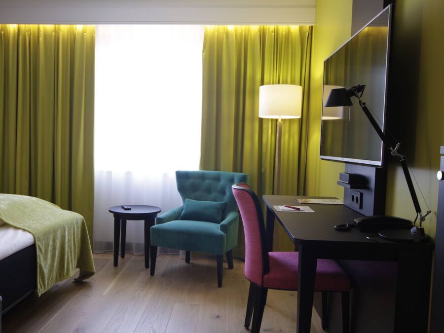 Quarto de hotel com cama, escrivaninha, cadeira, abajur e espelho.