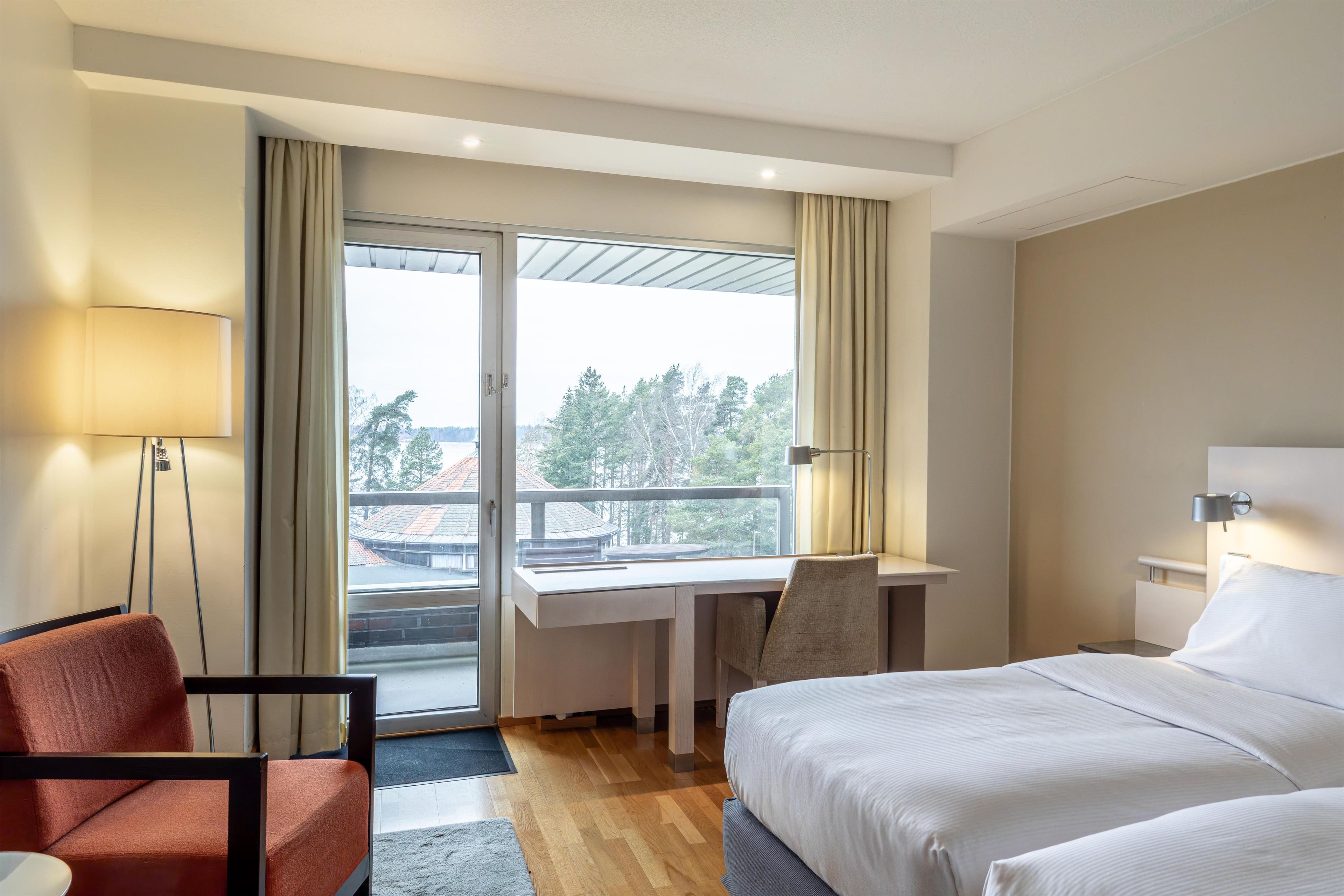 Quarto de hotel com cama, escrivaninha, cadeira e vista para as árvores pela janela.