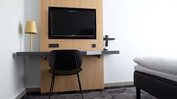 Quarto de hotel compacto com escrivaninha de madeira, uma cadeira preta, televisão na parede, mesa de cabeceira e cama com roupa de cama branca.
