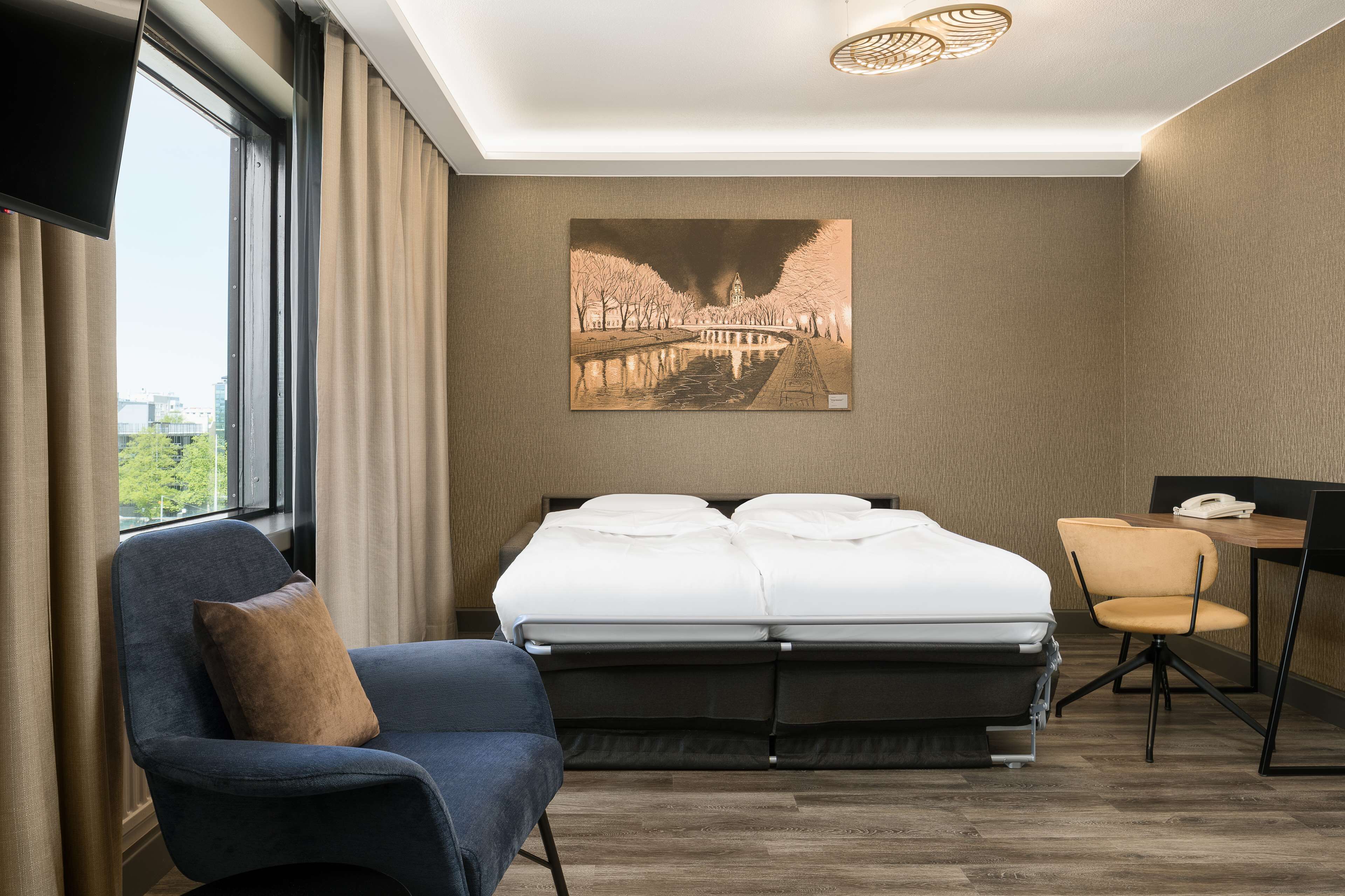 Quarto de hotel com cama, poltrona, escrivaninha e um quadro na parede.