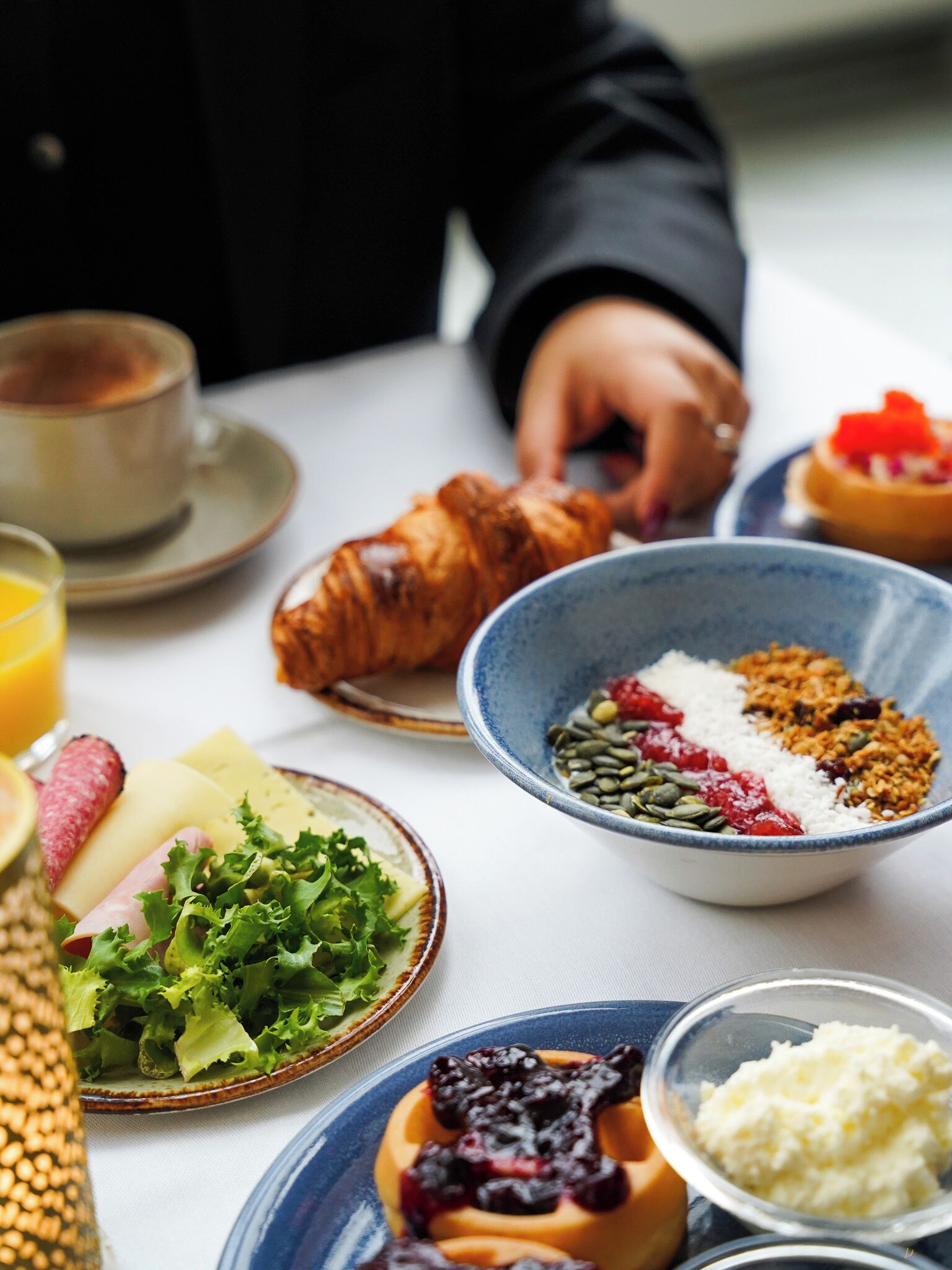 Uma mesa com diversos itens de café da manhã, incluindo croissants, uma tigela de cereal com frutas e sementes, um prato de vegetais e um copo de suco de laranja.