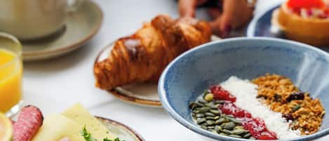 Uma mesa com diversos itens de café da manhã, incluindo croissants, uma tigela de cereal com frutas e sementes, um prato de vegetais e um copo de suco de laranja.