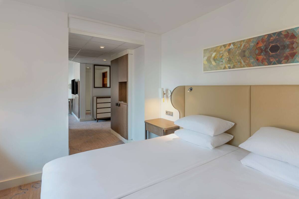 Quarto de hotel com cama, mesa de cabeceira, luminária na parede e quadro sobre a cama.