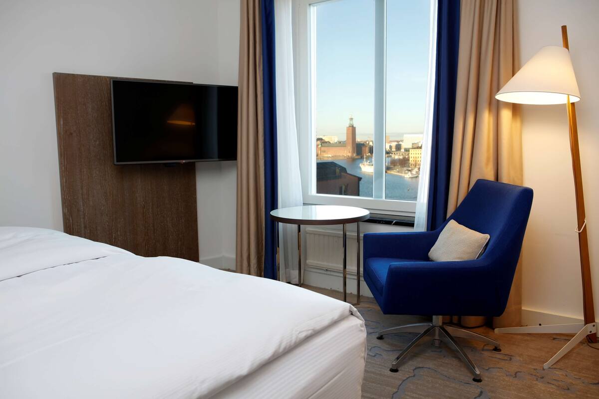 Quarto de hotel com cama, televisão fixada em painel de madeira, mesinha, poltrona azul, luminária e janela com vista para o horizonte da cidade.