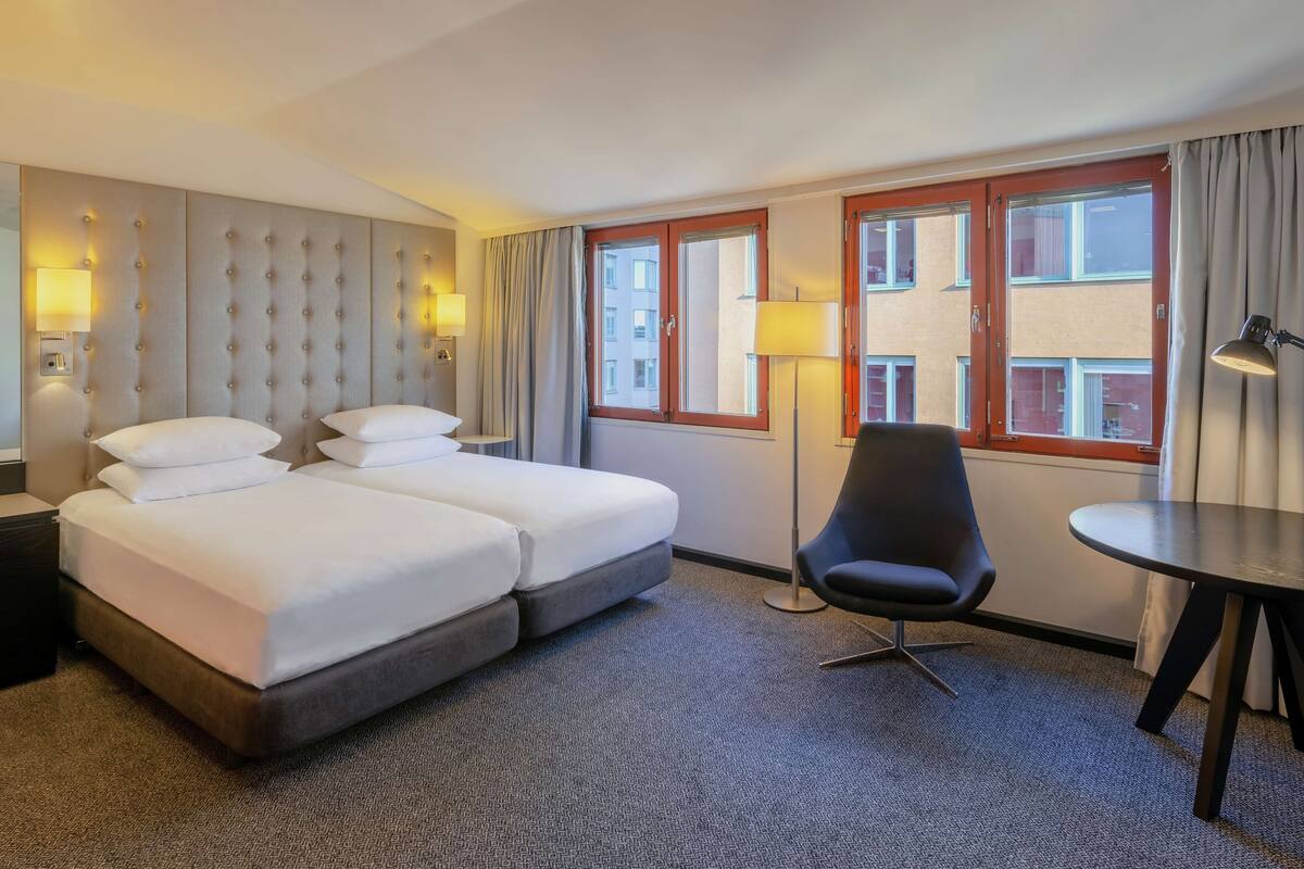 Quarto de hotel com duas camas, uma cadeira, uma mesa e uma janela com cortinas.