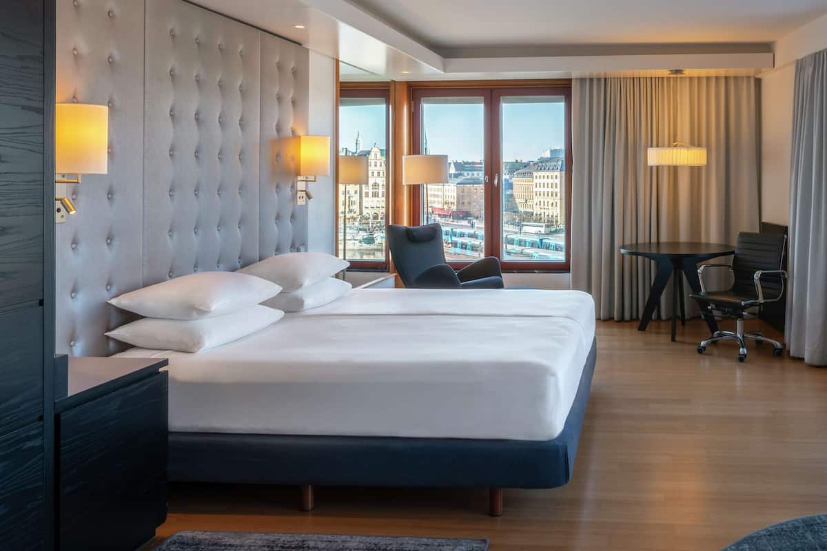 Quarto de hotel com uma cama grande, duas mesas de cabeceira, uma escrivaninha com cadeira e vista para a cidade através de grandes janelas.