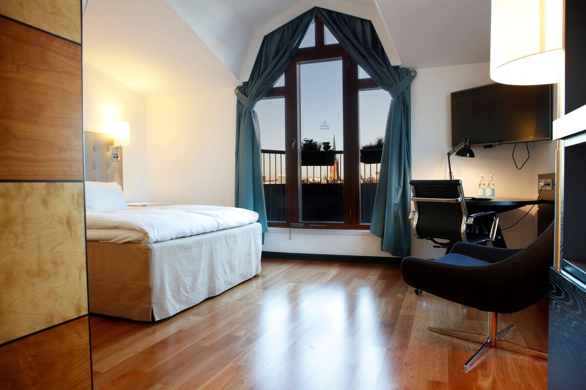Quarto de hotel com uma cama, uma escrivaninha com cadeira, uma televisão e uma janela com cortinas.