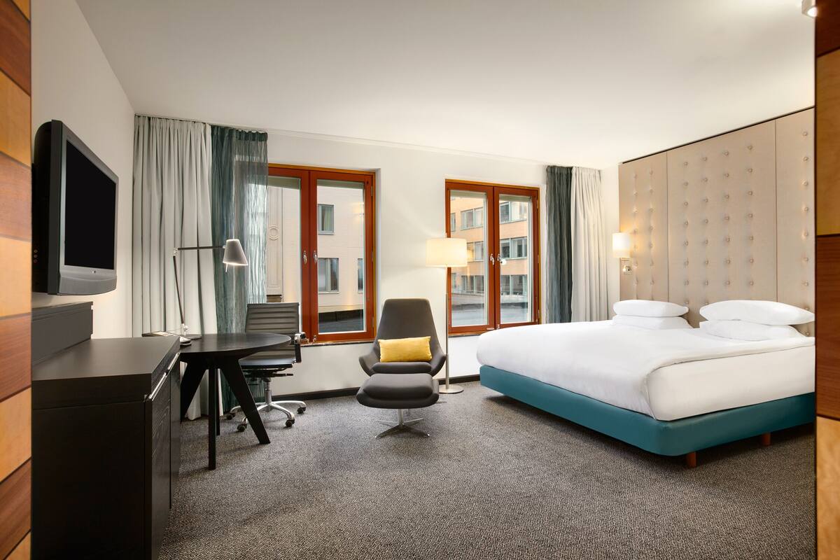 Quarto de hotel com cama, escrivaninha, cadeira, televisão e janela com cortinas.