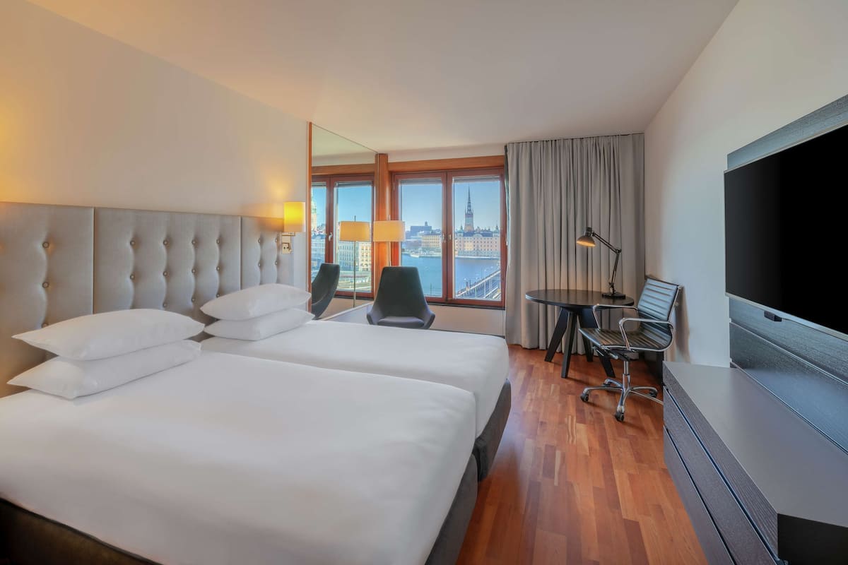 Quarto de hotel com duas camas, uma escrivaninha, uma cadeira, televisão e uma janela grande com vista para a cidade.