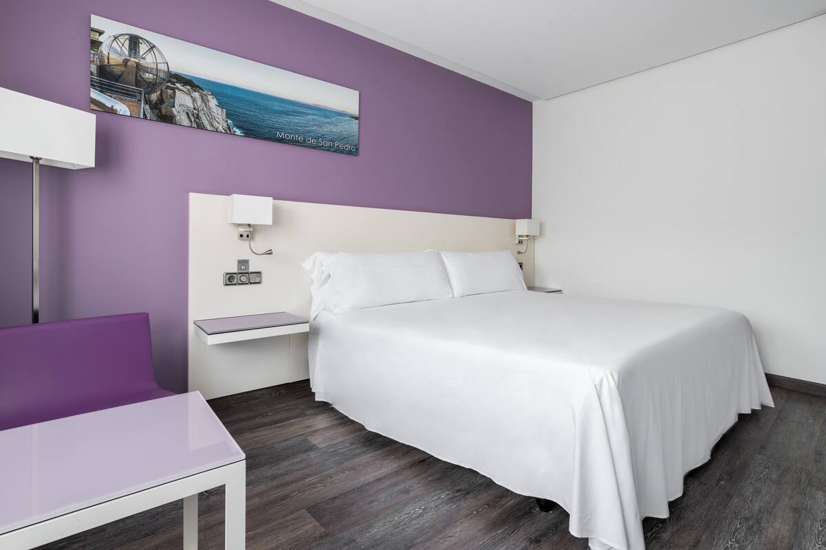 Quarto de hotel bem arrumado com uma cama, uma mesinha e uma cadeira. Há um quadro na parede retratando uma paisagem costeira.