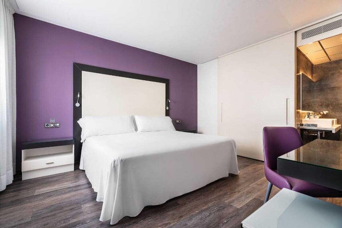 Quarto de hotel moderno com uma cama grande, uma secretária pequena e casa de banho visível por uma porta aberta.
