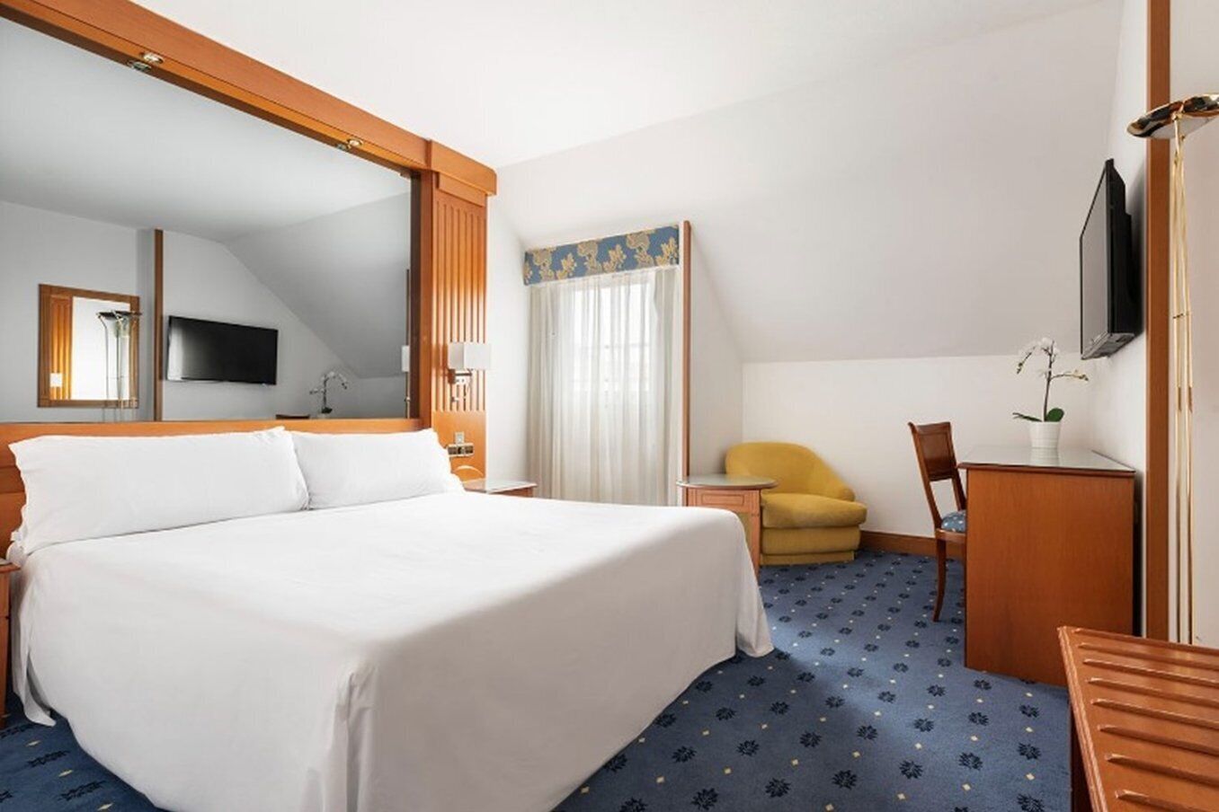 Quarto de hotel com uma cama grande, escrivaninha, cadeira, televisão e janela com cortinas.