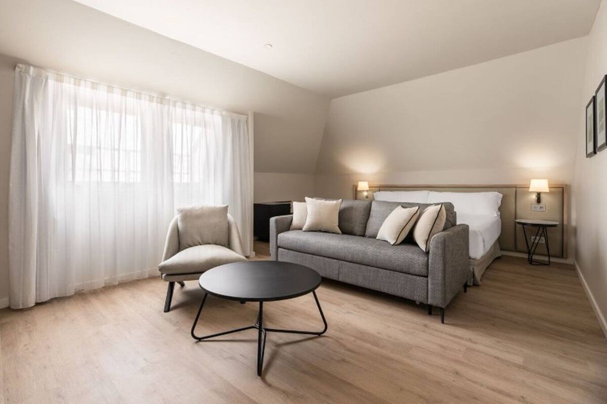 Quarto de hotel moderno com sofá cinza, mesa redonda preta, uma poltrona e uma cama com cabeceira. Há duas luminárias de cabeceira e uma mesinha com abajur.