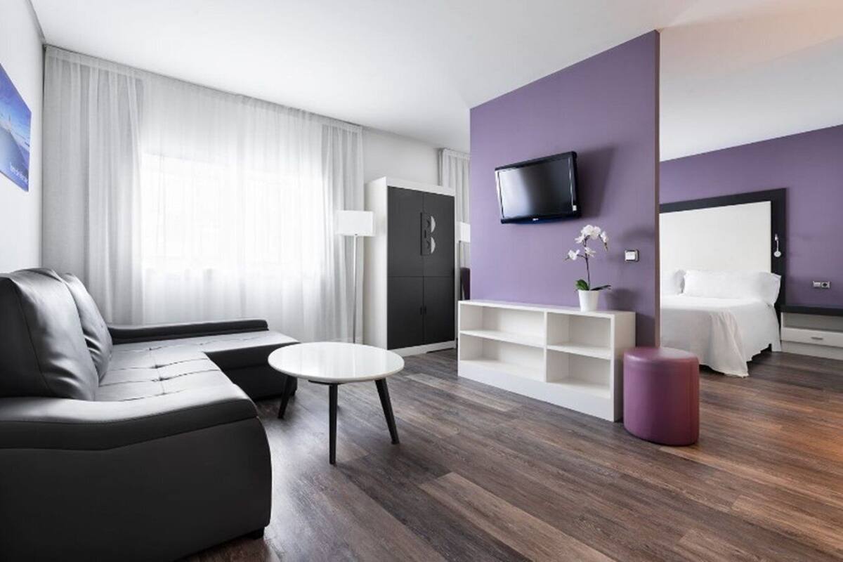 Quarto de hotel moderno com sofá cinza, mesa redonda branca, banquinho roxo, TV fixada na parede e cama com roupa de cama branca.