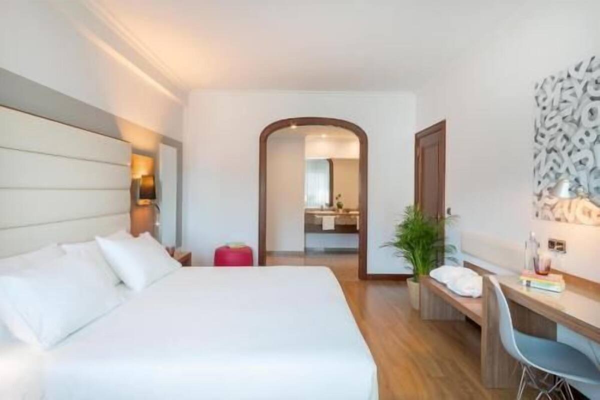 Quarto de hotel com uma cama grande, uma escrivaninha, uma cadeira, uma mesinha de centro, uma planta e um banheiro visível por um arco.