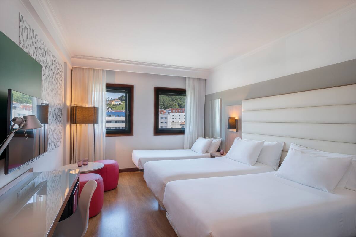 Quarto de hotel com duas camas, uma escrivaninha com televisão de tela plana, um banquinho vermelho e vista para prédios pelas janelas.