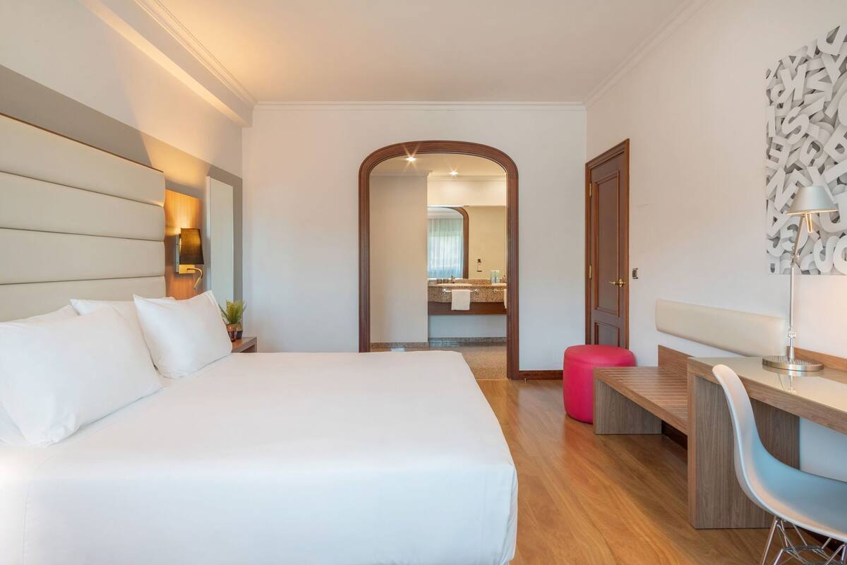 Quarto de hotel com uma cama grande, uma escrivaninha, uma cadeira, um banquinho pequeno, um espelho e um banheiro visível através de um arco.