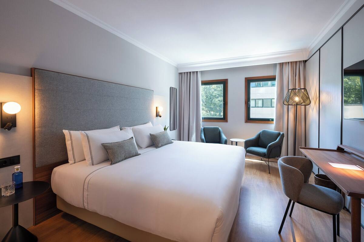 Quarto de hotel com uma cama grande, duas cadeiras, uma escrivaninha e um abajur.