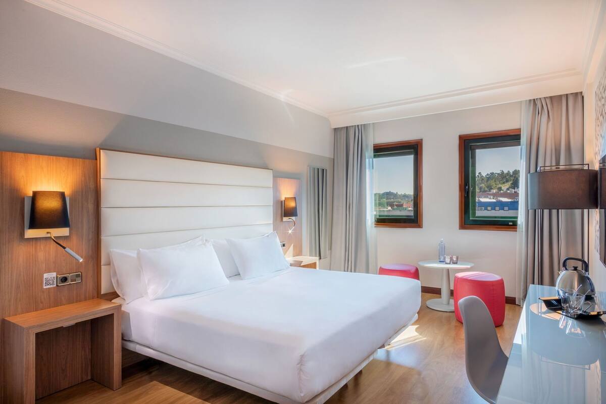 Quarto de hotel com uma cama grande, mesas de cabeceira, uma escrivaninha com cadeira, uma mesinha redonda e vista para o exterior.