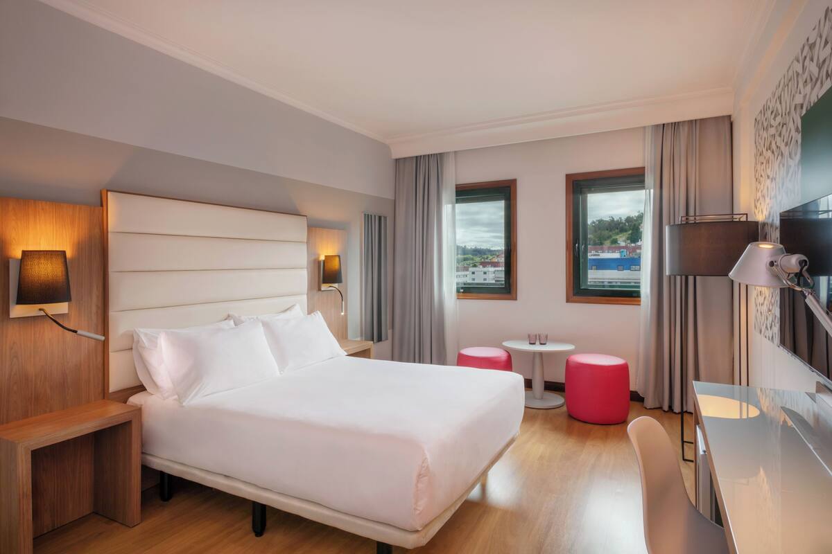 Quarto de hotel moderno com uma cama grande, mesas de cabeceira, uma escrivaninha, duas cadeiras e uma pequena mesa com um banquinho vermelho.