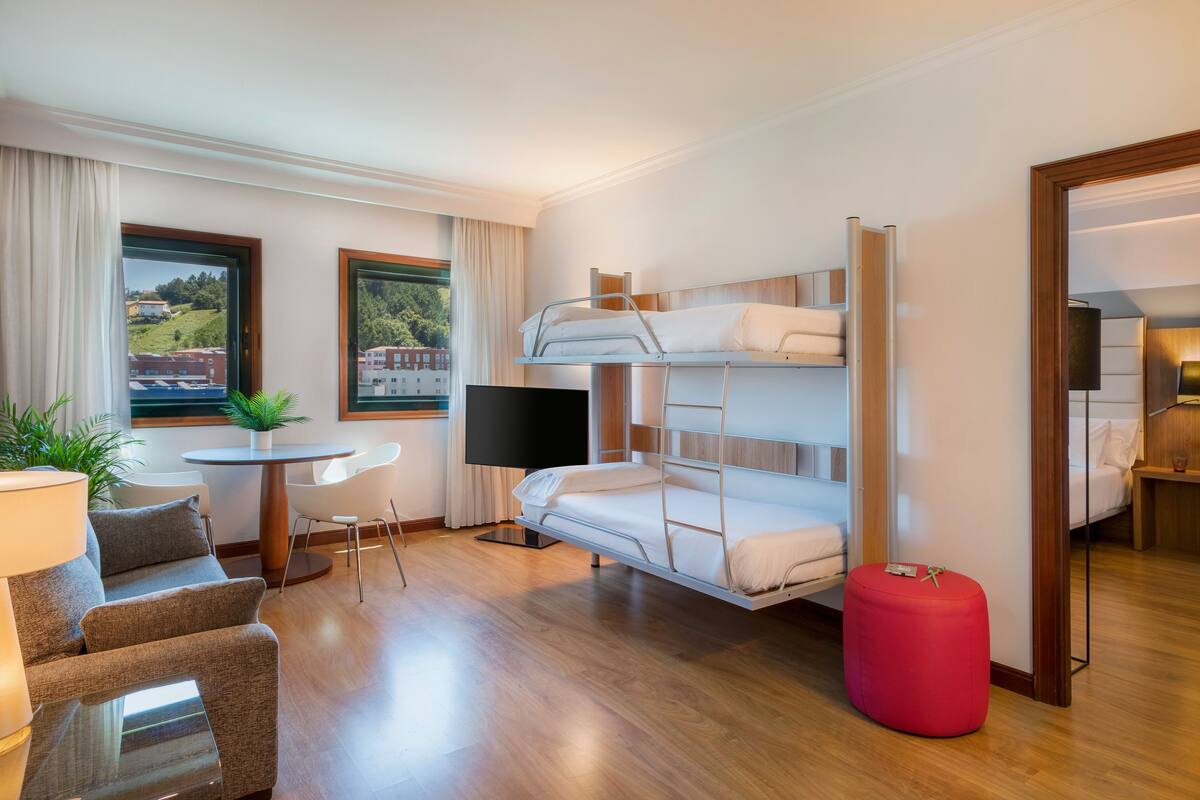 Quarto de hotel com beliche, uma mesa pequena com cadeiras, televisão e um banquinho vermelho.