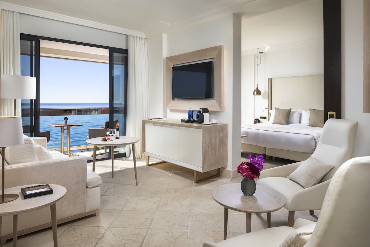 Quarto de hotel moderno com uma grande janela, televisão de tela plana, sofá, poltrona, mesa pequena e cama.