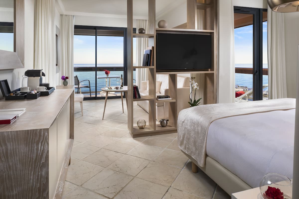 Quarto de hotel com uma cama grande, uma escrivaninha com um laptop, uma TV e uma varanda com vista para o mar.