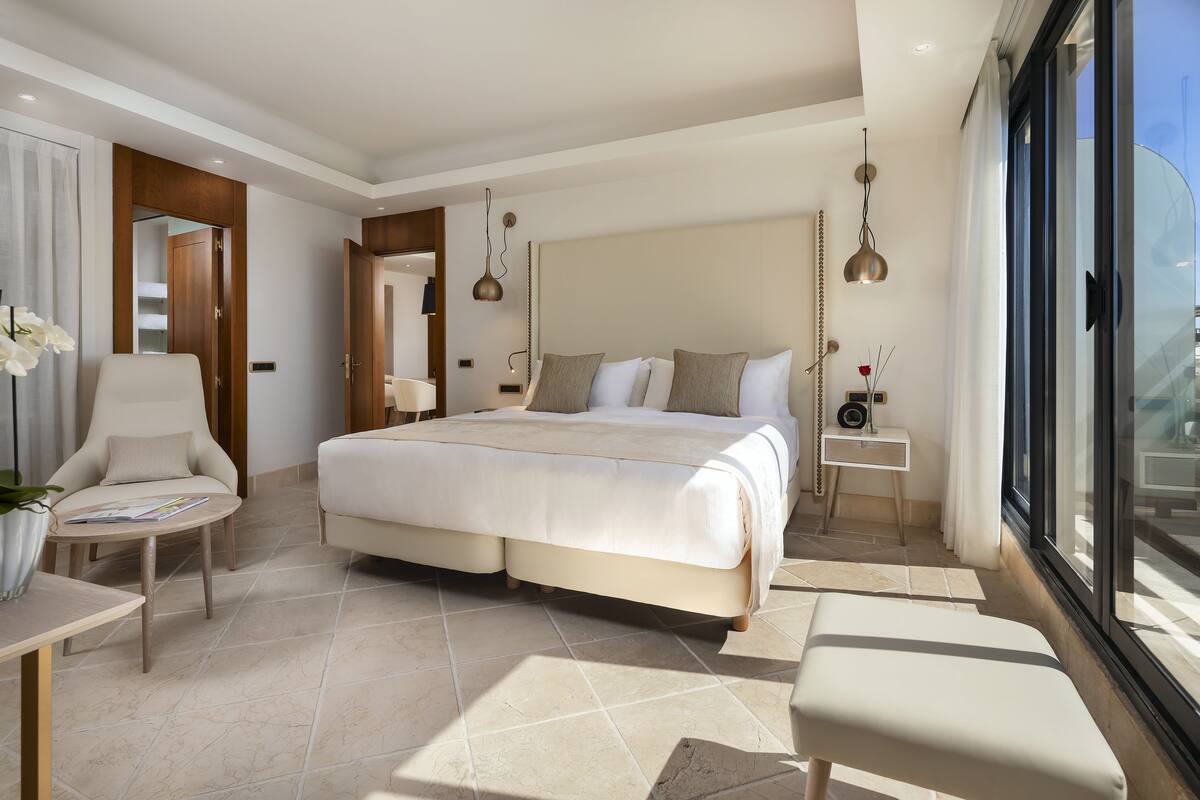 Quarto de hotel moderno com uma cama grande, um espaço de estar com uma poltrona e mesa, e vista pela janela ampla.