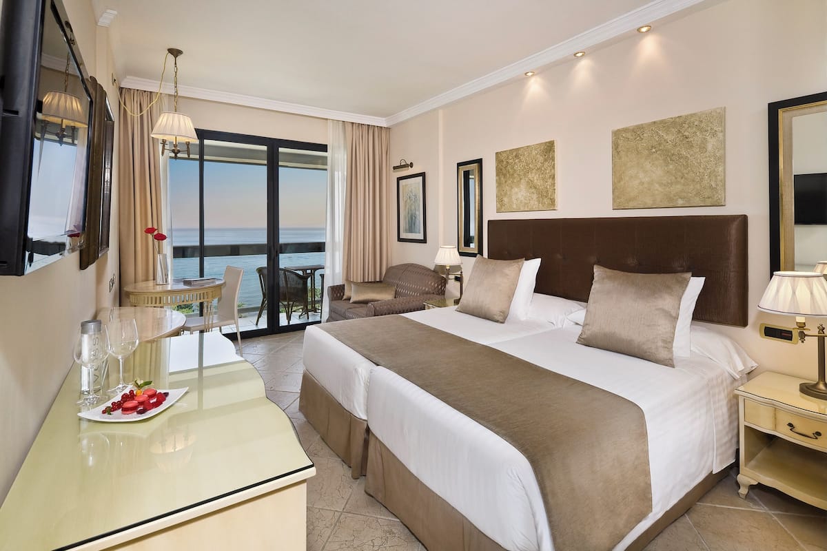 Quarto de hotel com uma cama grande, uma mesa de jantar com cadeiras, varanda com vista para o mar e uma televisão na parede.