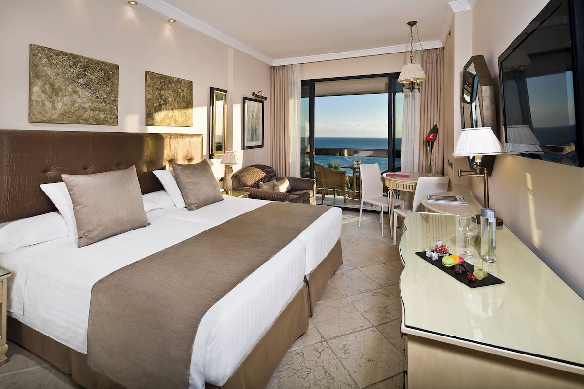 Quarto de hotel com uma cama grande, um espaço de estar com mesa e cadeiras, e uma varanda com vista para o mar.
