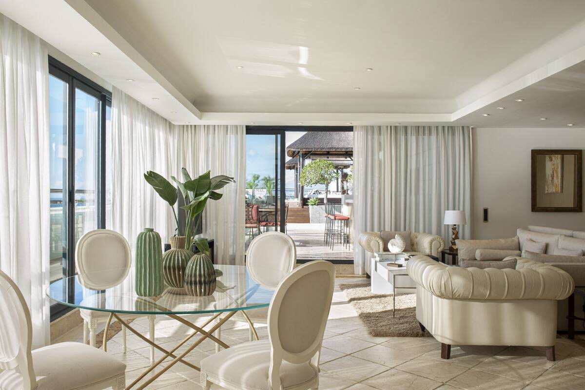 Uma sala de estar espaçosa com uma mesa de jantar de vidro, cadeiras brancas, um sofá bege e vista para a área externa.